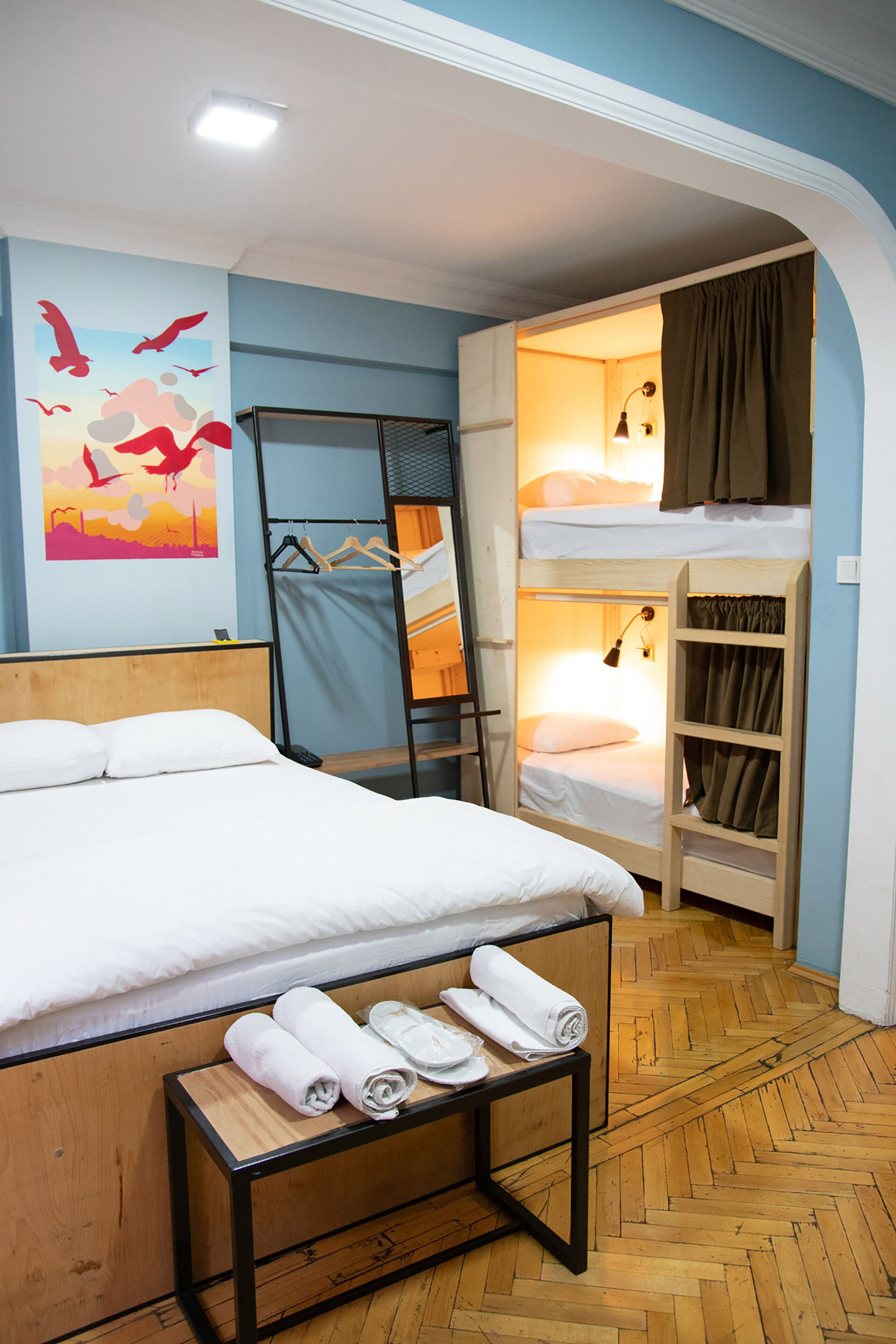 Quarto com uma cama de solteiro, uma cama individual, uma mesa de madeira com toalhas, uma arte na parede com pássaros e um chão com padrão espinha de peixe.