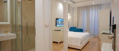 Quarto de hotel moderno com chuveiro em cabine de vidro, cama branca, escrivaninha com cadeira e televisão na parede.