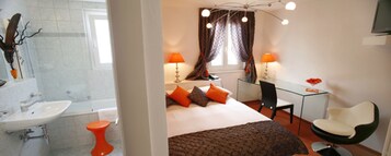 Quarto de hotel com cama, escrivaninha, cadeira, mesinha de vidro, banquinho vermelho, pia branca e banheira.