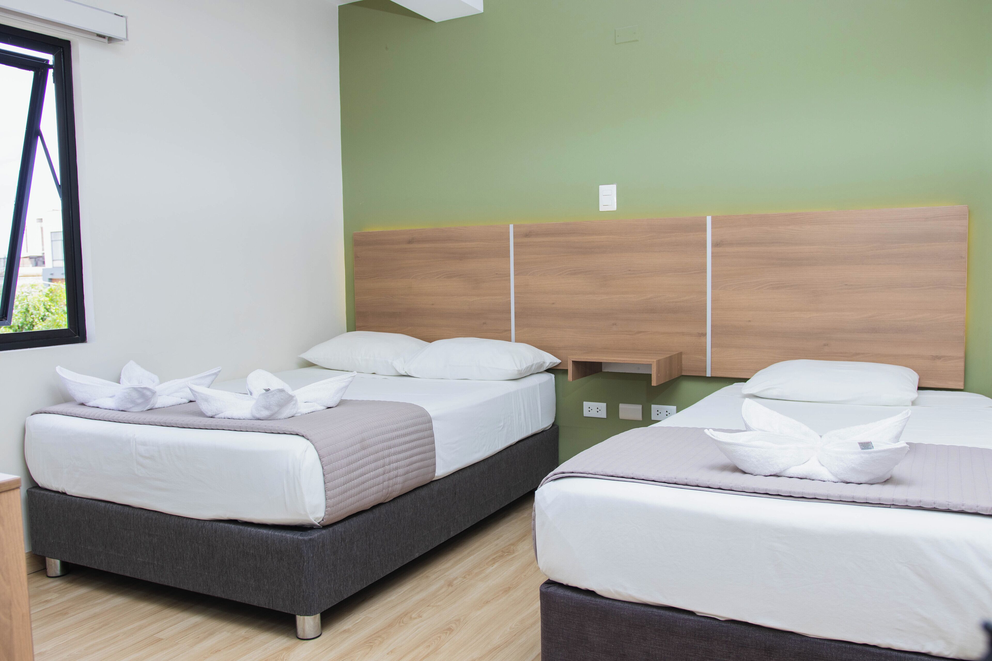 Quarto de hotel com duas camas, cada uma com roupa de cama branca e almofadas decorativas. O quarto tem uma parede verde e uma cabeceira de madeira.