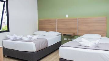 Quarto de hotel com duas camas, cada uma com roupa de cama branca e almofadas decorativas. O quarto tem uma parede verde e uma cabeceira de madeira.