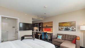 Quarto de hotel moderno com uma cama grande, um sofá, uma mesa com um computador, uma TV de tela plana e um banheiro visível por uma porta aberta.