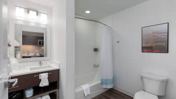 Um banheiro moderno com pia branca, espelho e vaso sanitário. Há um chuveiro com porta de vidro e cortina branca. Uma citação emoldurada está na parede.