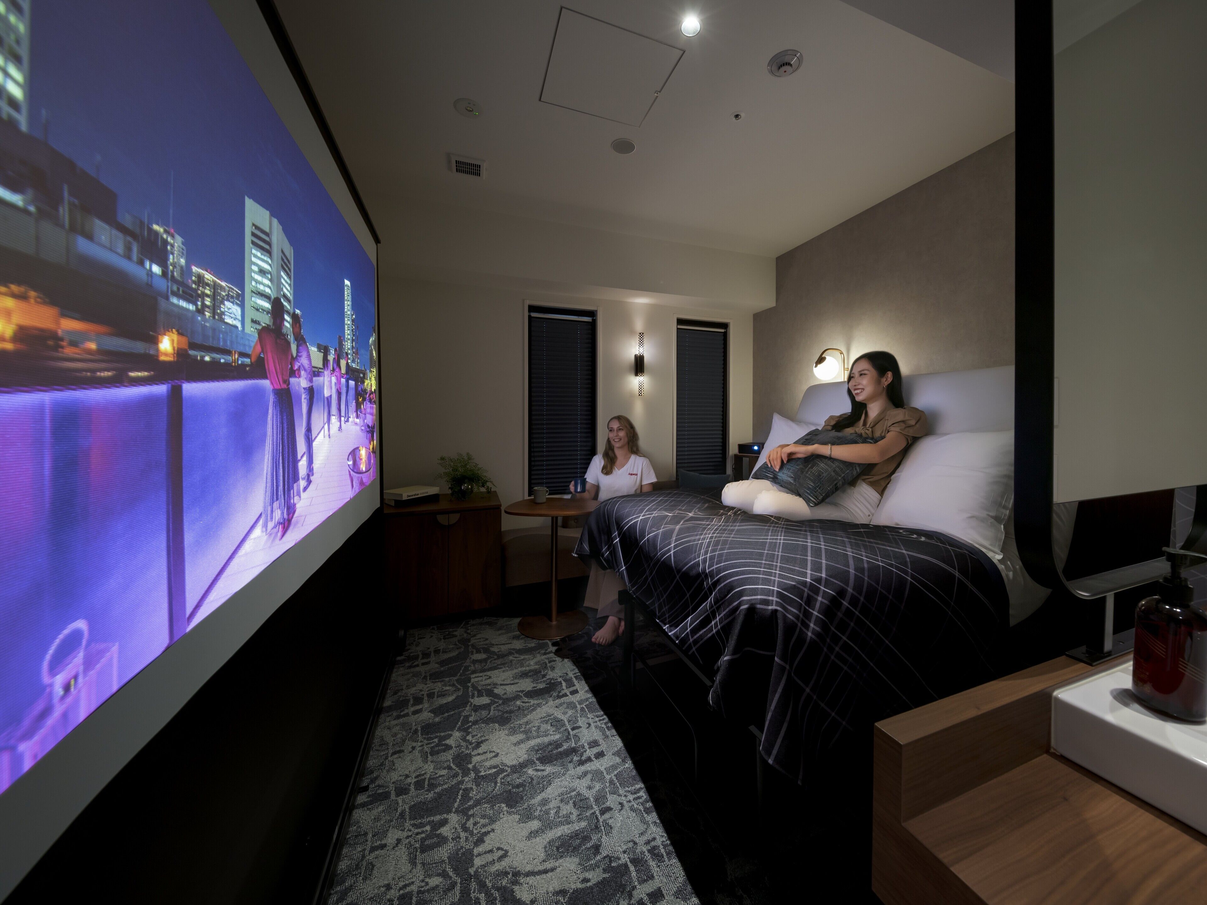 Quarto de hotel com uma tela grande exibindo uma vista urbana, cama com colcha xadrez, mesa de cabeceira com abajur e pequena mesa com uma planta e uma xícara.