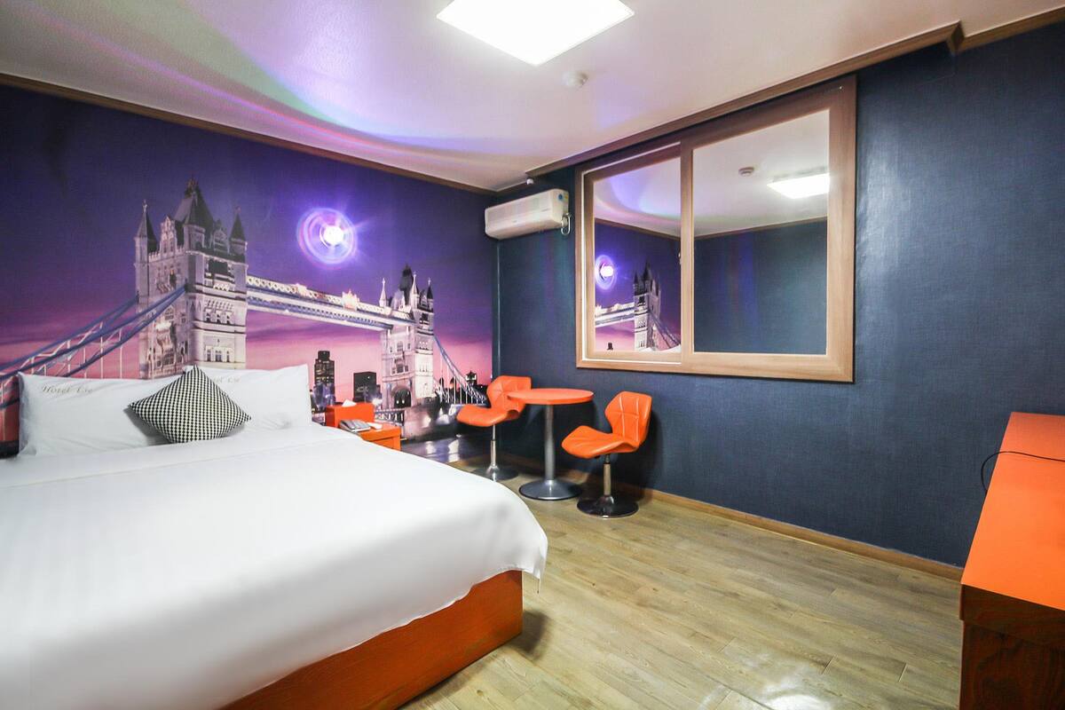 Quarto de hotel com cama, escrivaninha, duas cadeiras, espelho e um mural na parede retratando uma ponte.