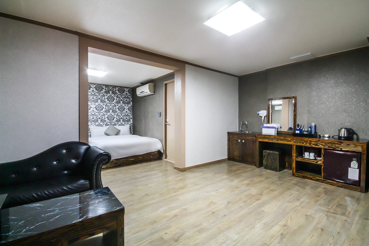 Quarto de hotel com uma cama, uma toucador de madeira, um espelho, uma poltrona de couro preto e piso de madeira clara.