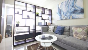 Uma sala de estar moderna com um sofá cinza, uma mesa de centro redonda de vidro e uma estante com moldura preta e diversos objetos decorativos.