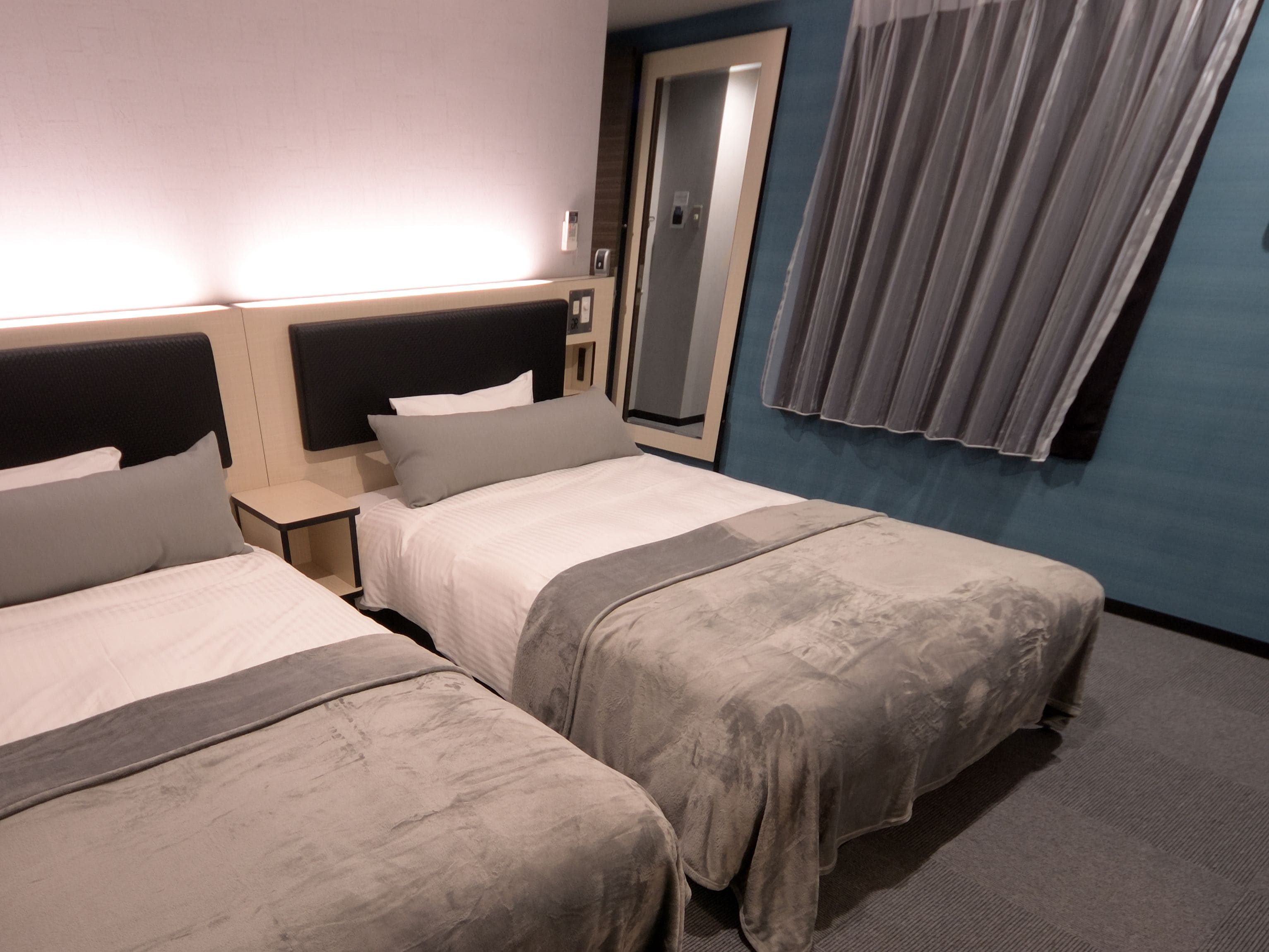 Quarto de hotel com duas camas, mesa de cabeceira, espelho e janela com cortinas.