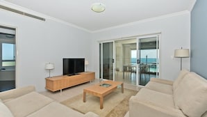 Sala de estar moderna com televisão de tela plana, móveis de madeira e porta de vidro que dá para uma varanda com vista para o mar.