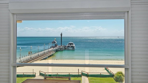 Vista da praia com um píer que se estende para o oceano, um farol e um barco pequeno.