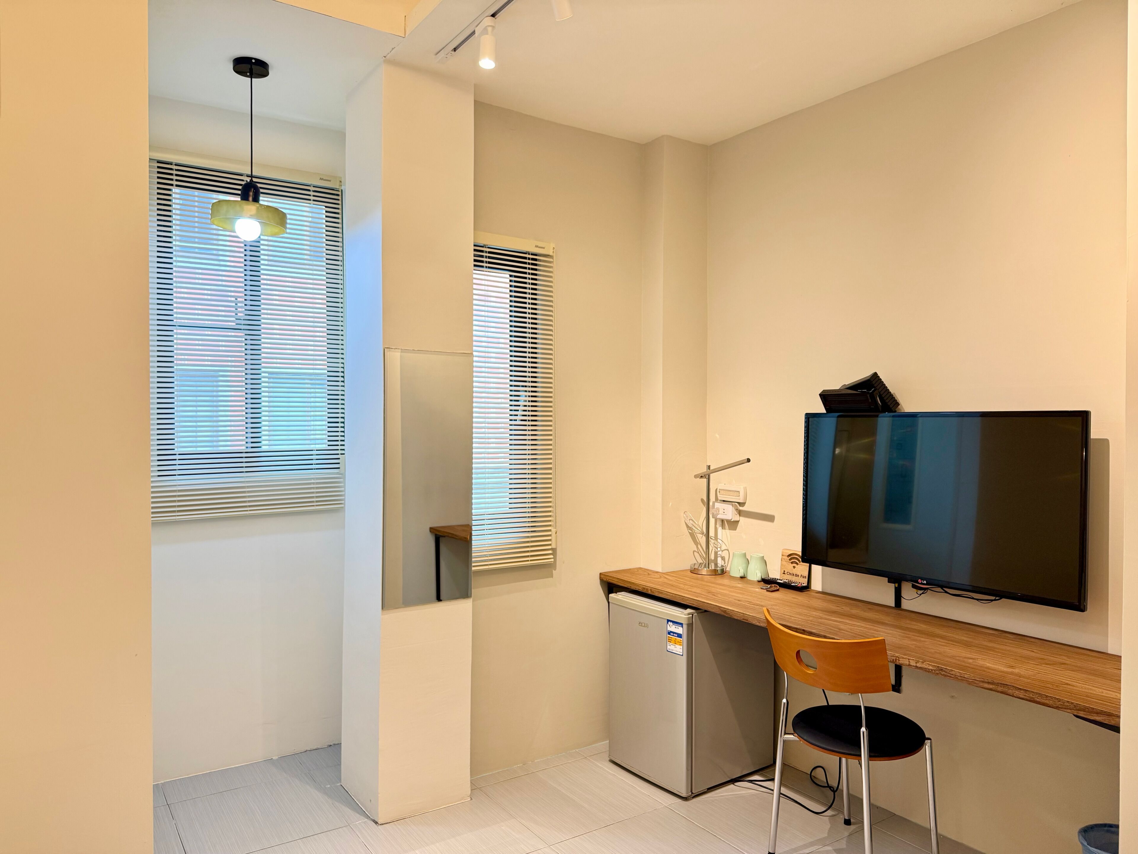 Quarto compacto com cozinha compacta, uma escrivaninha com cadeira, televisão e janela com persianas.