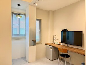 Quarto compacto com cozinha compacta, uma escrivaninha com cadeira, televisão e janela com persianas.