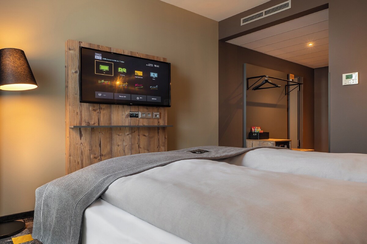 Quarto de hotel com uma cama, televisão presa a um painel de madeira, luminária de chão e espelho.