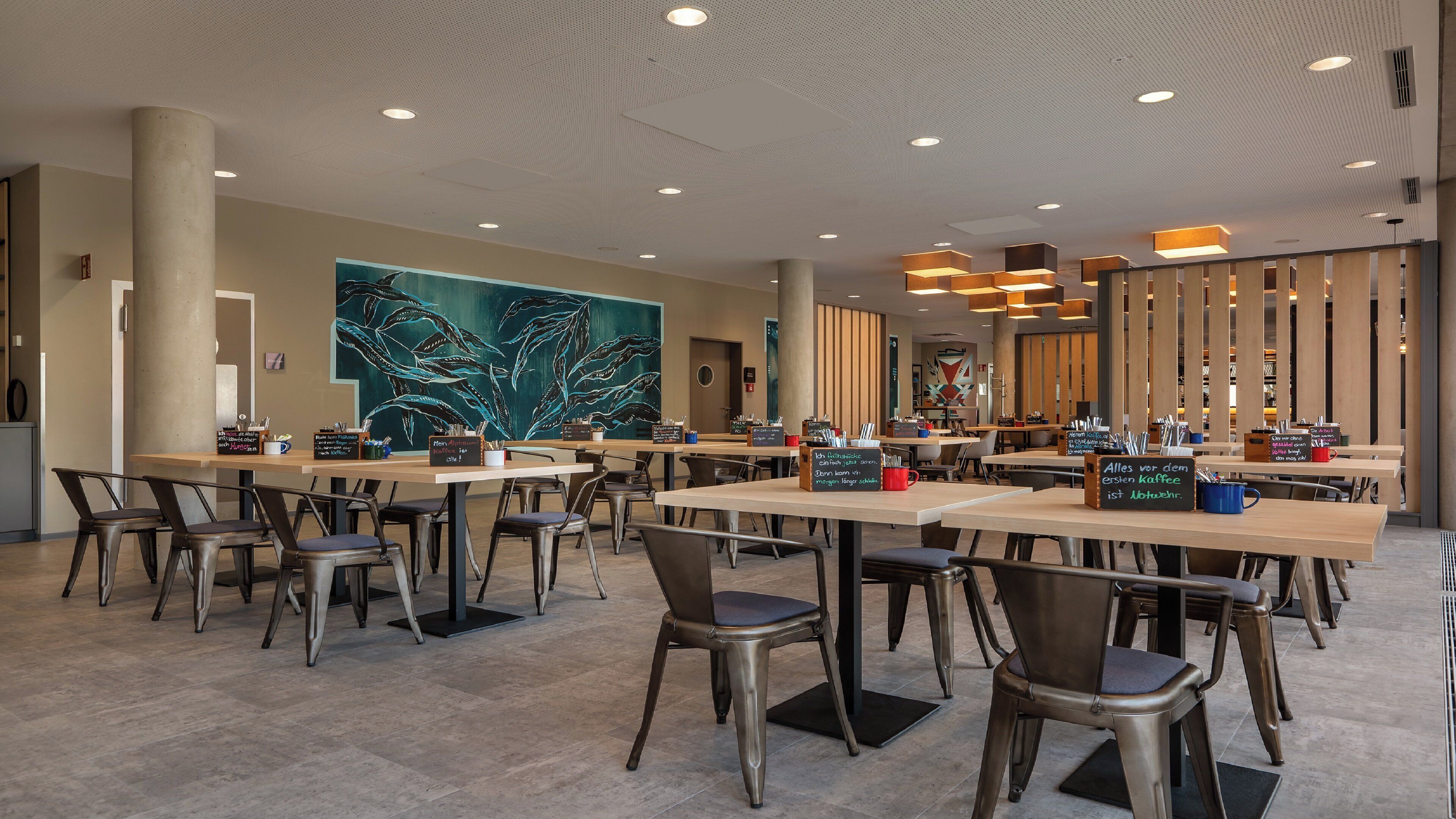 Uma ampla sala de jantar com várias mesas e cadeiras, um grande quadro abstrato na parede e luminárias modernas.