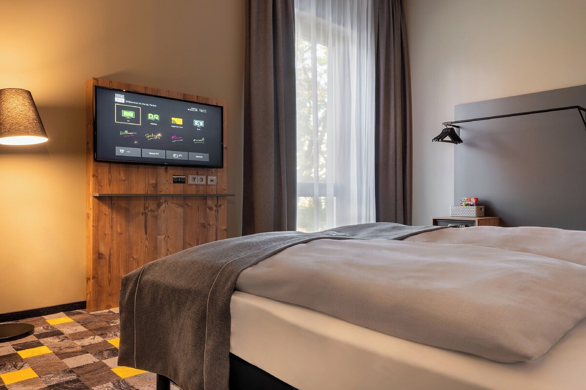 Quarto de hotel moderno com cama, televisão de tela plana fixada em um painel de madeira, abajur e mesa de cabeceira com luminária.