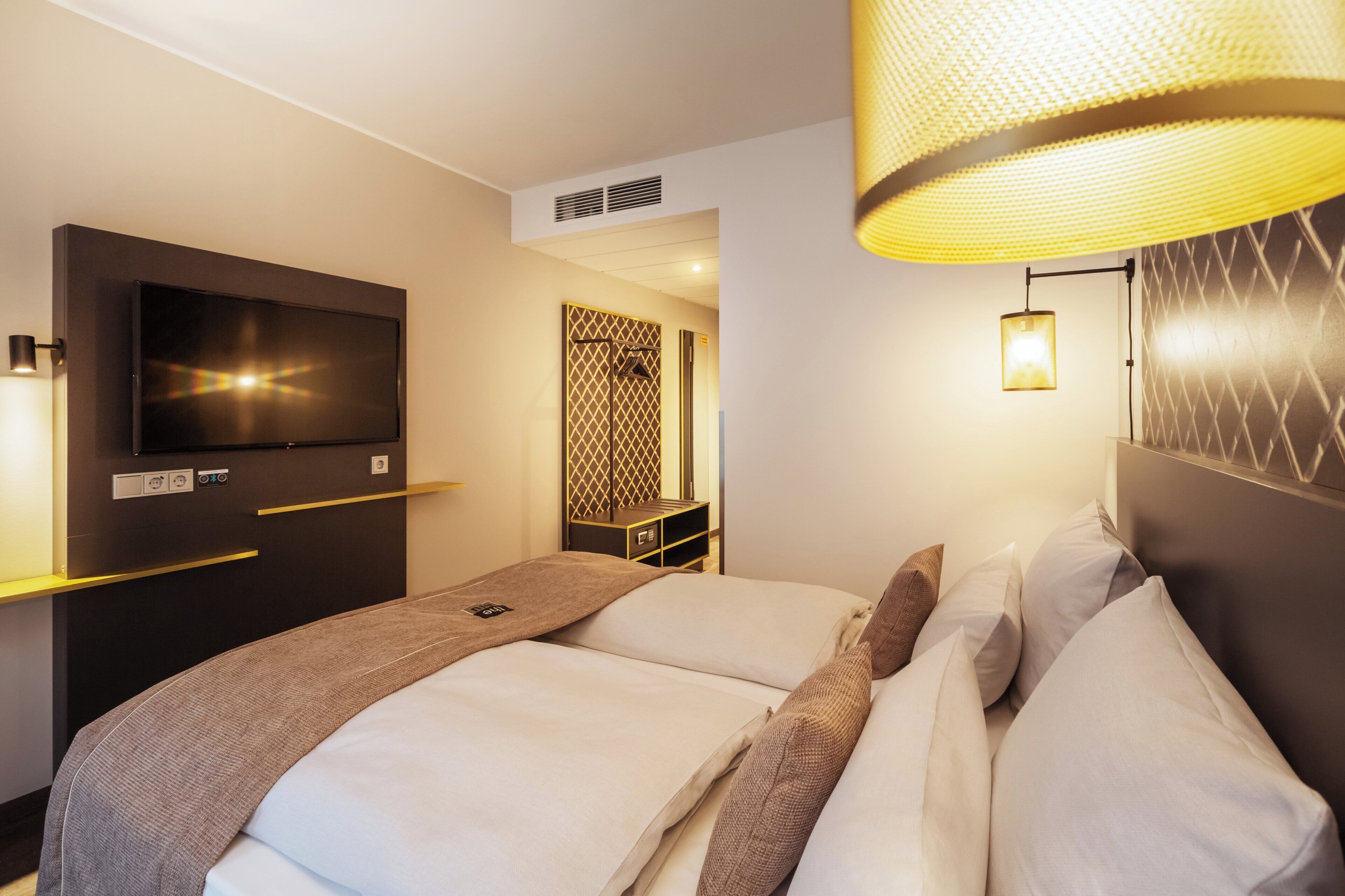 Quarto de hotel com uma cama grande, televisão de tela plana, televisão fixada na parede e uma luminária moderna.