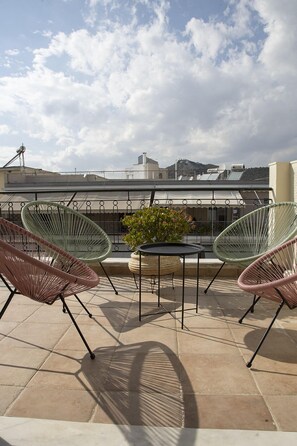 Terraço no telhado com cadeiras de vime e uma mesa, um vaso de planta e vista de prédios ao fundo.