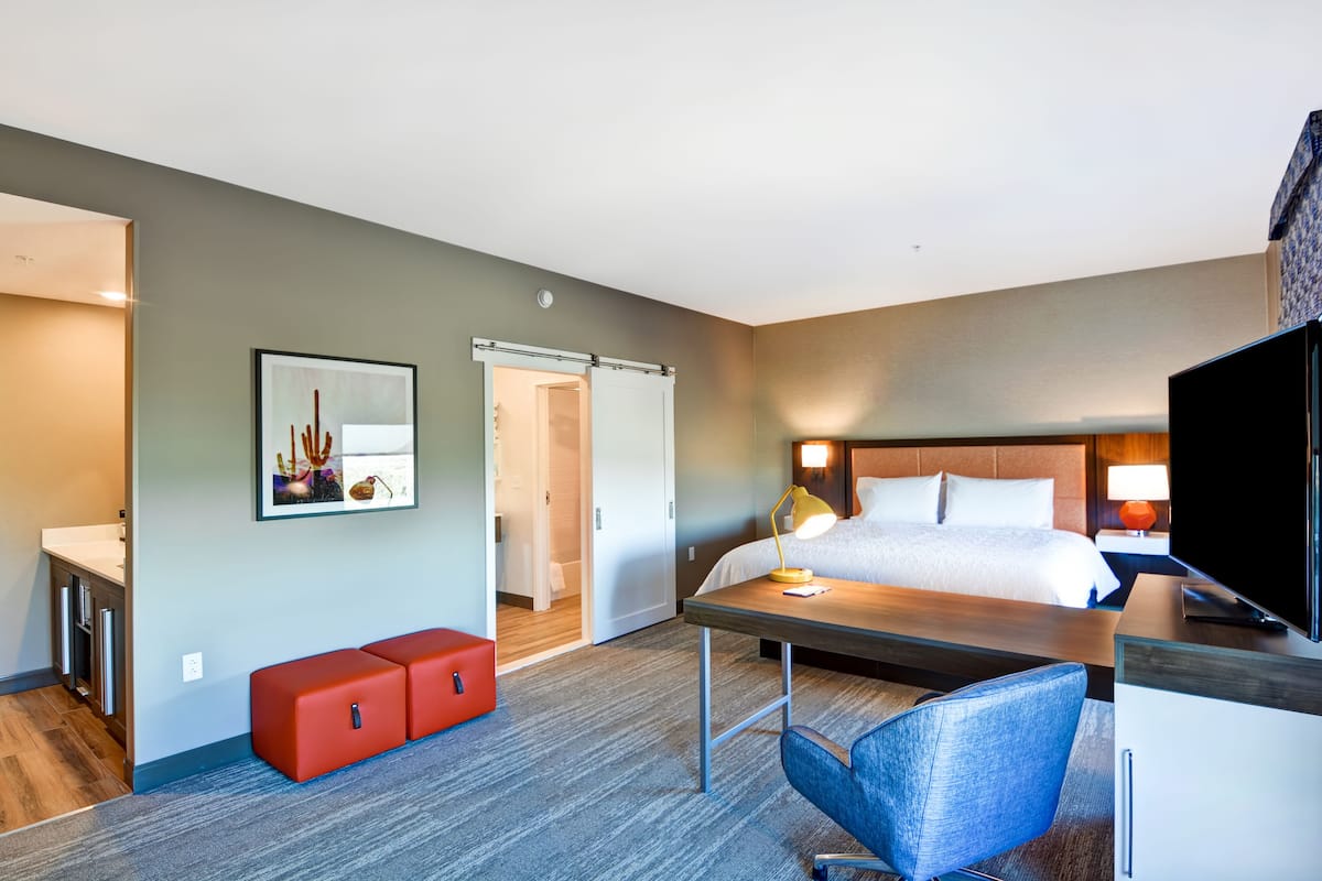 Quarto de hotel com cama, escrivaninha, cadeira, televisão, um banco vermelho e porta para o banheiro.