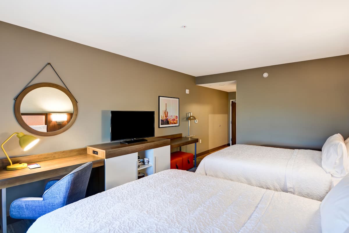 Quarto de hotel com duas camas, uma mesa de trabalho com cadeira, televisão e espelho.
