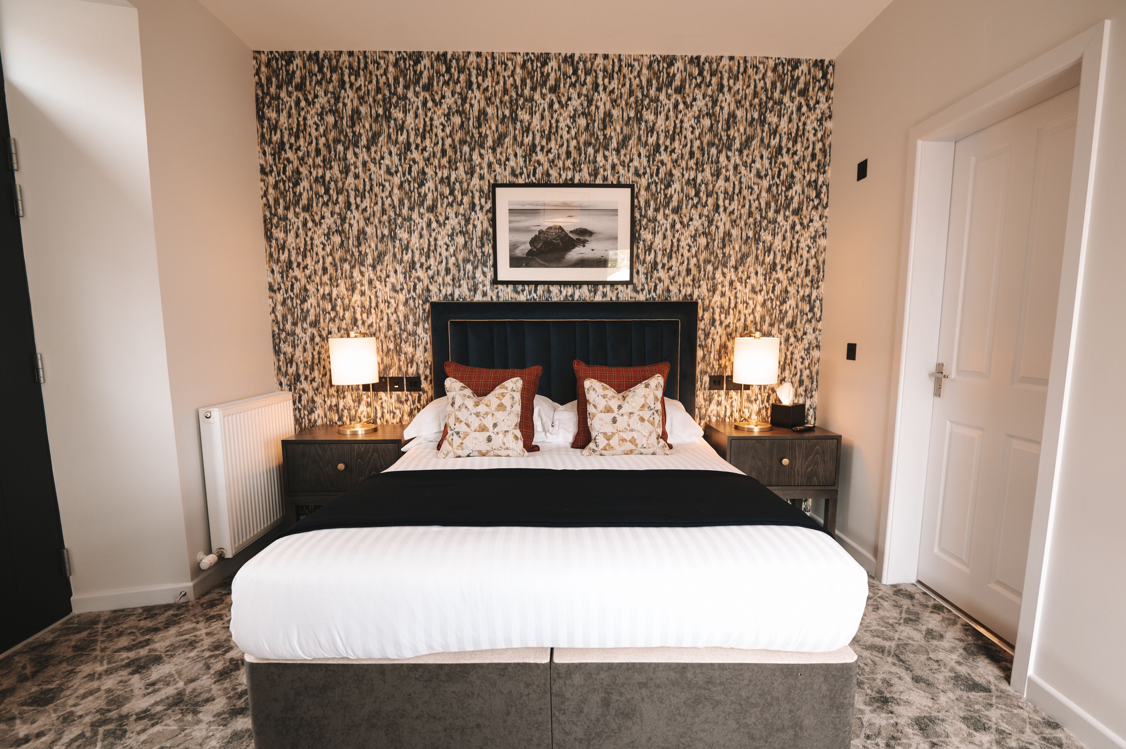 Cama bem arrumada com cabeceira escura, duas luminárias de cabeceira e um quadro emoldurado acima da cama.