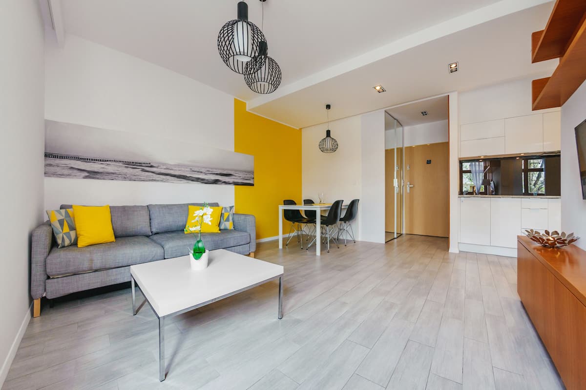 Uma sala de estar moderna com sofá cinza, almofadas amarelas, mesa de centro branca e um espaço de jantar com mesa e cadeiras.
