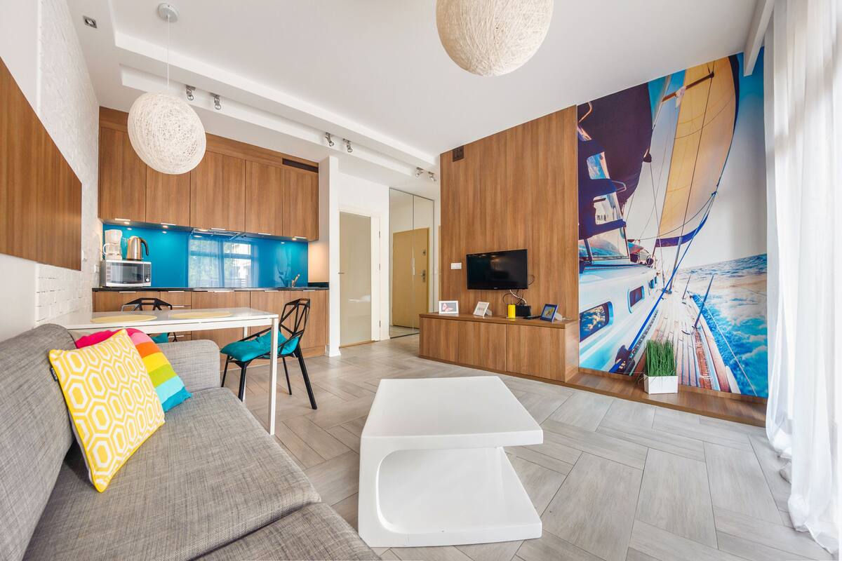Uma sala de estar moderna com um sofá cinza, almofadas coloridas, uma mesa de centro branca e um mural de um veleiro na parede.