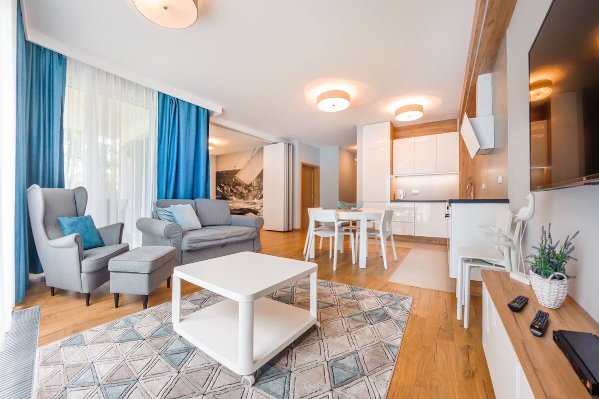 Uma sala de estar moderna com sofá, apoio para os pés e mesa de centro, área de jantar com cadeiras, cozinha com armários brancos e uma janela grande com cortinas azuis.
