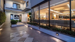 Exterior de hotel moderno com fachada de vidro, área de estar externa e entrada bem iluminada.