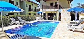 Uma piscina com espreguiçadeiras e guarda-sóis, rodeada por um pátio de pedra e um edifício com varandas.