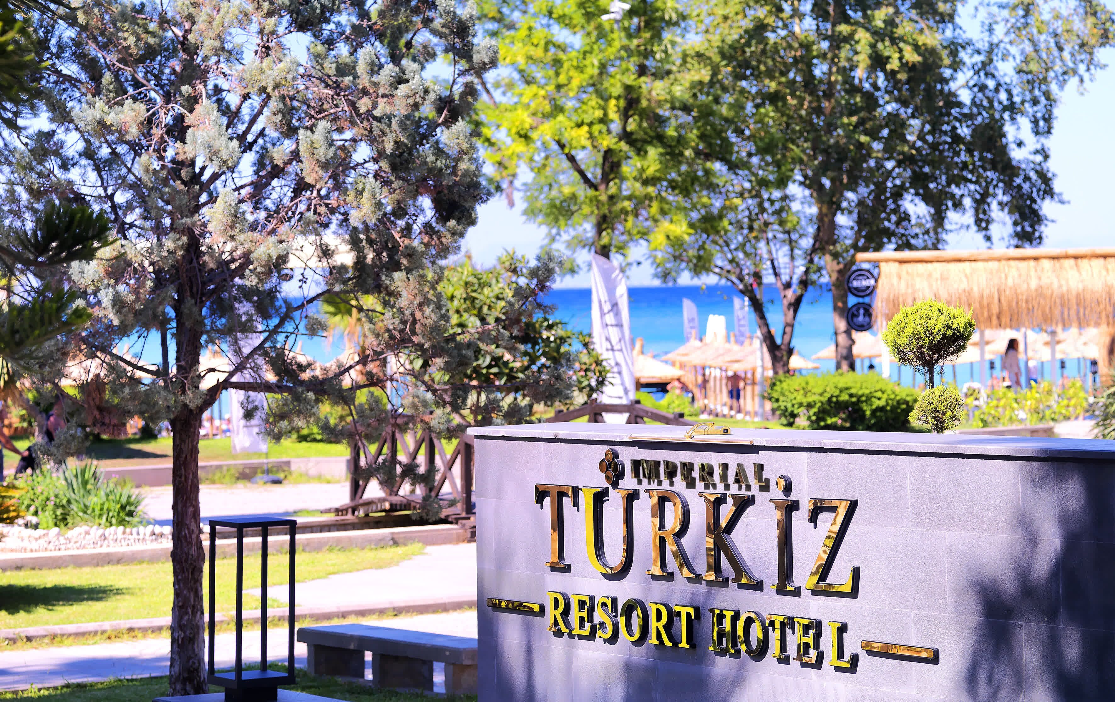 Exterior do Hotel Imperial Turkish Resort, com uma placa, cercado por árvores e vista para o mar.