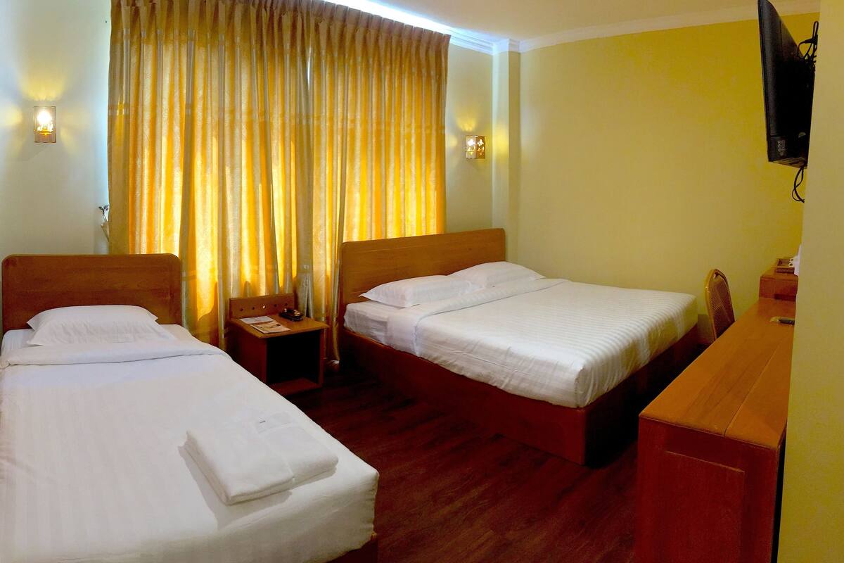 Quarto de hotel com duas camas de solteiro, cabeceiras de madeira, escrivaninha de madeira, uma cadeira, televisão fixada na parede e cortinas.