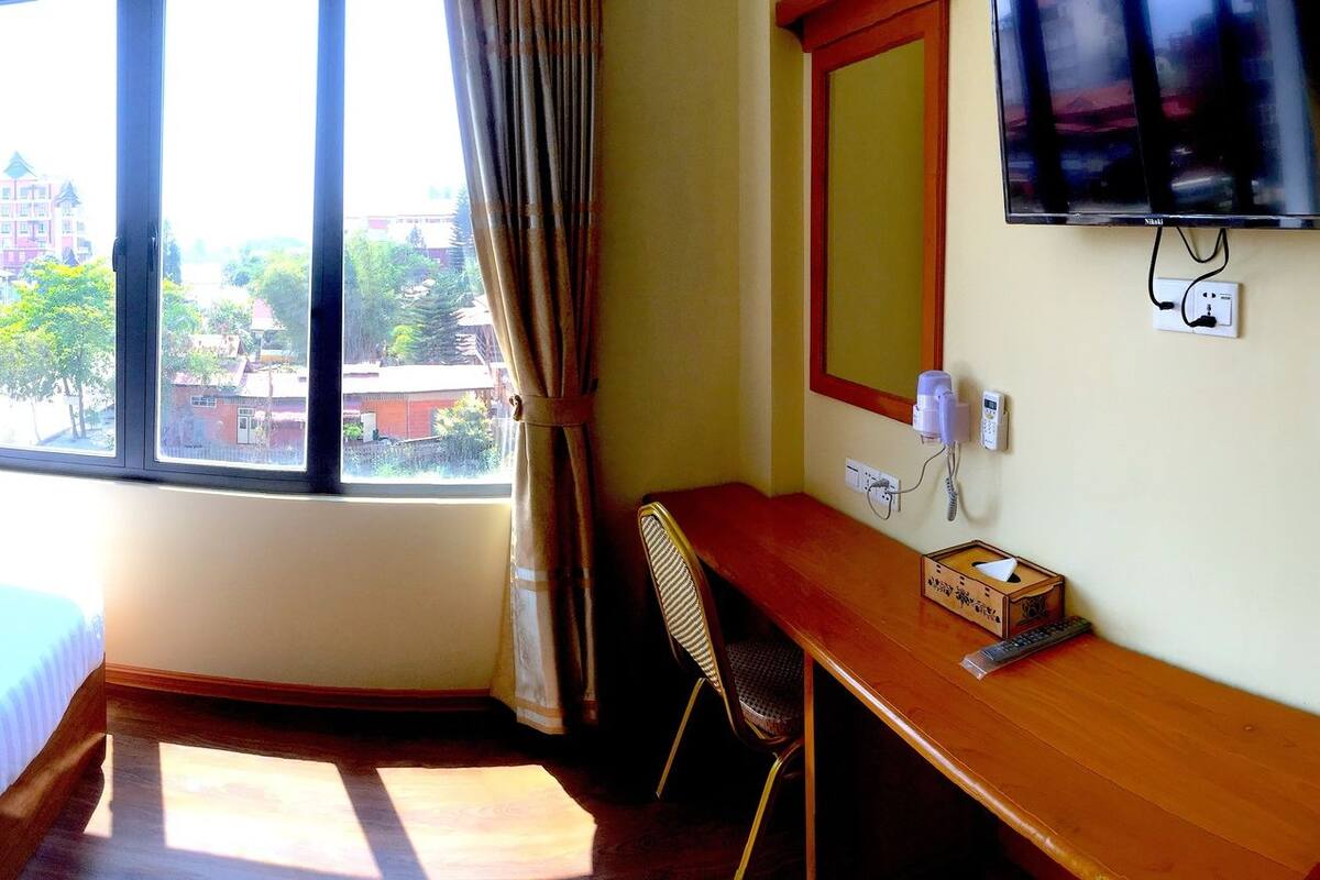 Quarto de hotel com cama, escrivaninha, cadeira, televisão e janela com vista para edifícios.