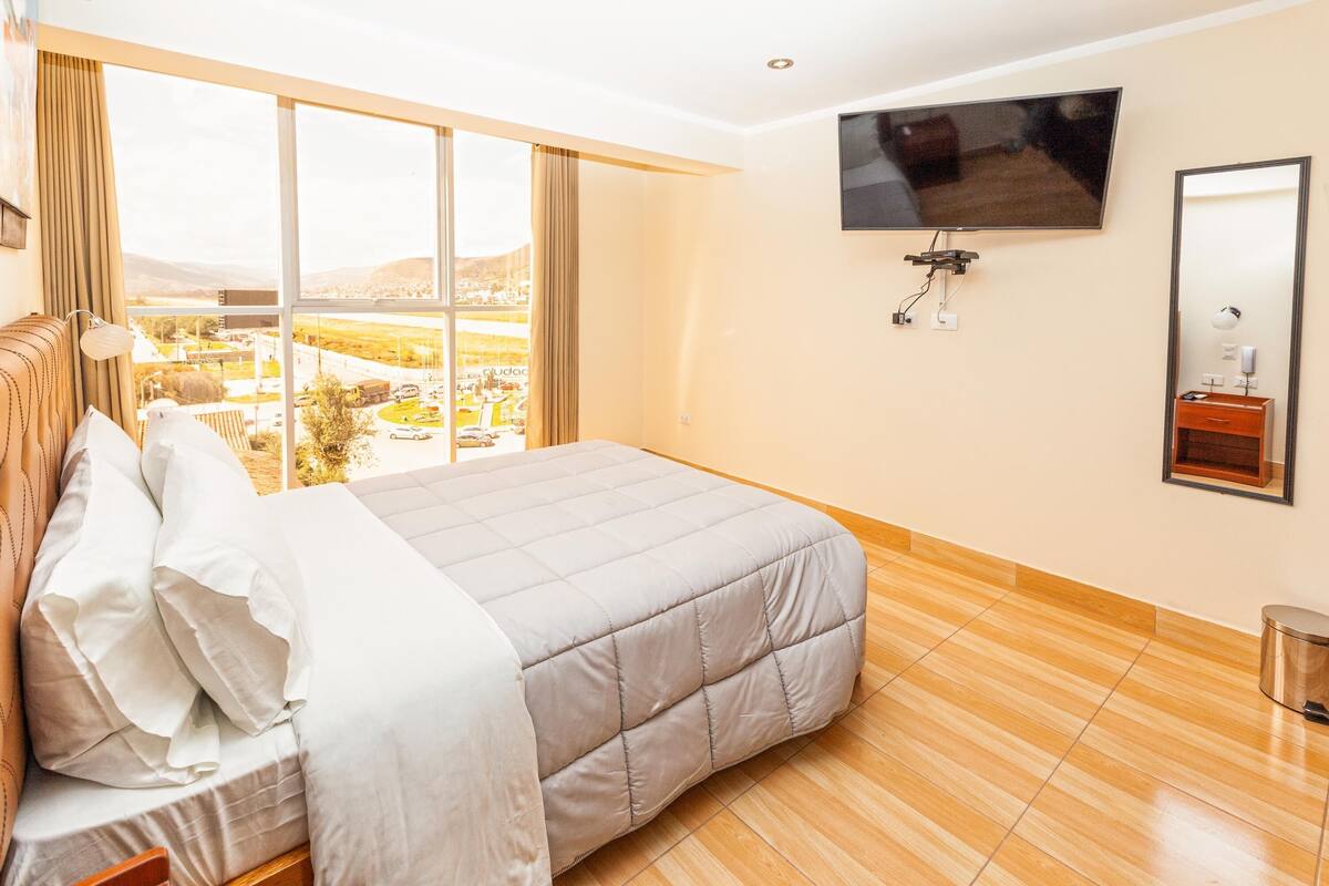 Quarto de hotel com uma cama grande, televisão fixada na parede, espelho e vista para a paisagem urbana através de amplas janelas.