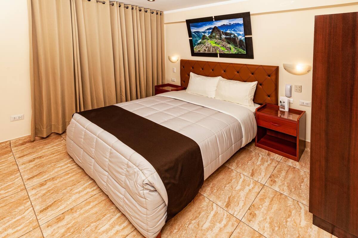 Quarto de hotel com cama, mesa de cabeceira, televisão fixada na parede e guarda-roupa de madeira.