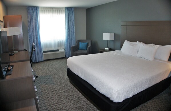 Quarto de hotel com uma cama grande, uma escrivaninha com luminária, uma cadeira e uma janela com cortinas.