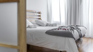 Uma cama bem arrumada com lençóis brancos e um cobertor cinzento, uma cabeceira de madeira e uma janela com cortinas translúcidas.