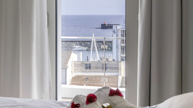 Quarto de hotel com uma cama, janela com cortinas e vista para o mar e edifícios.