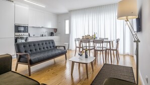 Uma sala de estar moderna com sofá, mesa de centro e um espaço de jantar com cadeiras e mesa. Há também uma cozinha com armários e eletrodomésticos brancos.