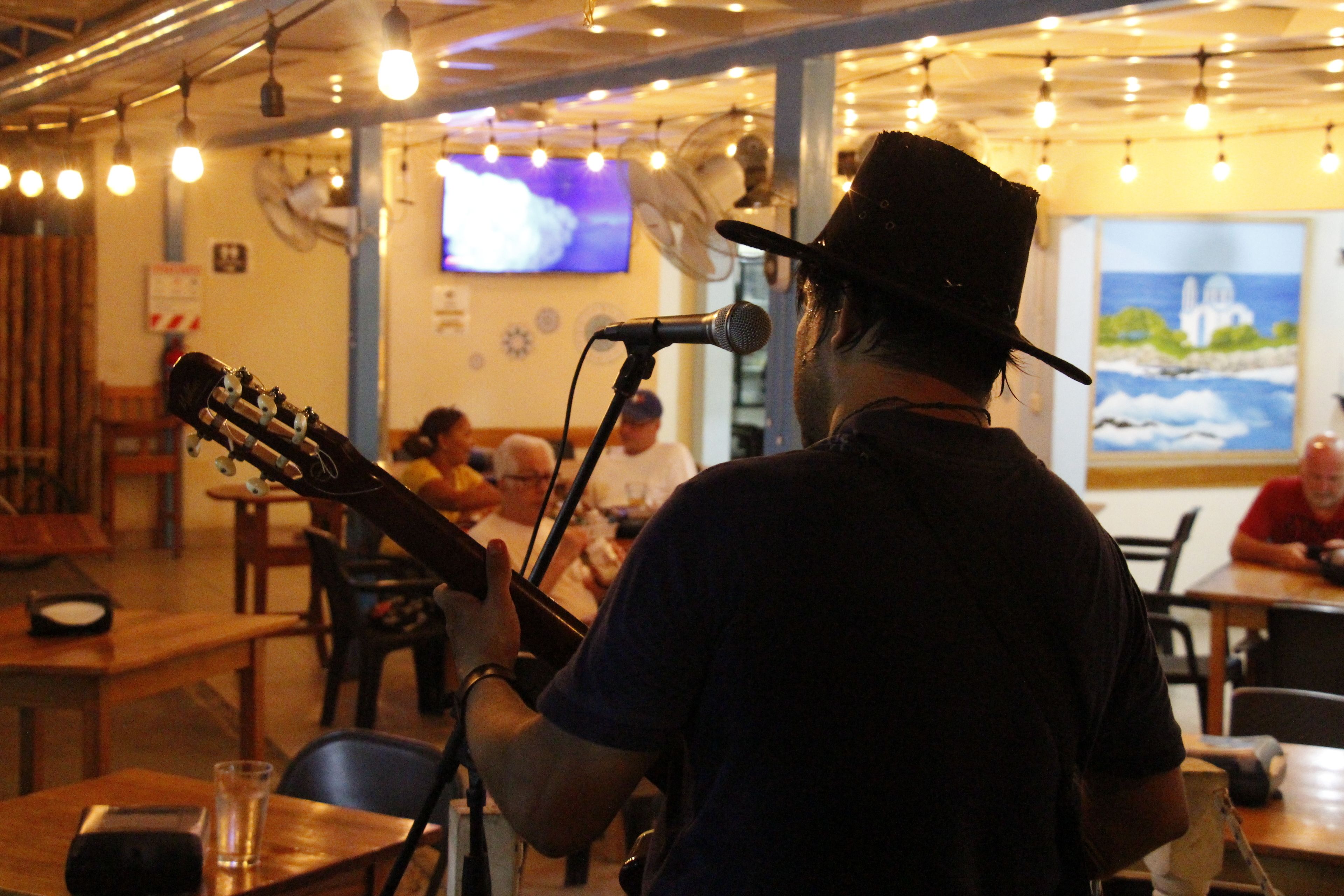 Uma pessoa tocando violão em um microfone em restaurante, com mesas e cadeiras, uma televisão na parede e um quadro retratando uma paisagem costeira.