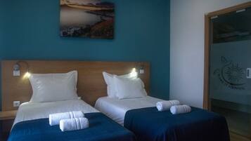Quarto de hotel com duas camas, cada uma com lençóis brancos e colchas azuis. Há um quadro com um pôr do sol na parede, e um logotipo na porta.