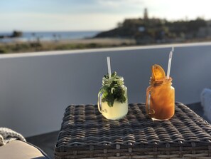 Duas bebidas sobre uma mesa de vime, com vista para a praia e um edifício distante.