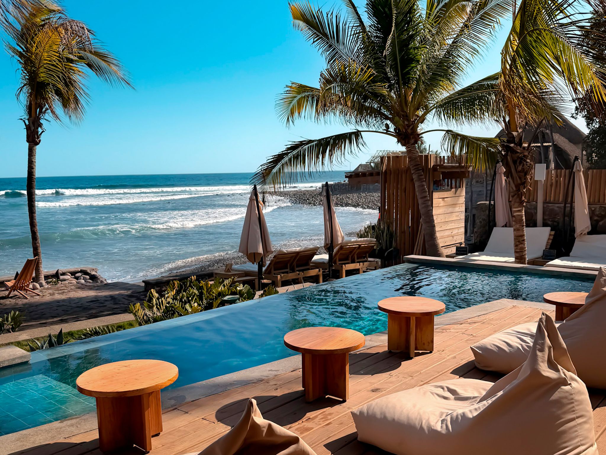 Área de piscina com espreguiçadeiras e guarda-sóis, com vista para uma praia e oceano.