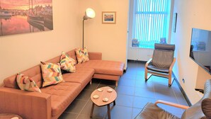 Sala de estar com sofá, mesa de centro, poltrona, televisão, quadro, abajur e janela com persianas.