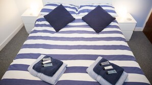 Cama arrumada com roupa de cama listrada e duas almofadas, duas toalhas dobradas e duas luminárias de cabeceira em mesas de cabeceira brancas.