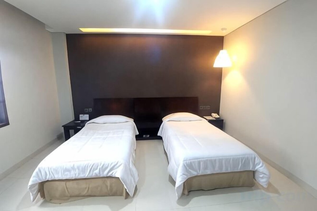 Quarto de hotel com duas camas, cabeceira escura, mesas de cabeceira e uma luminária na parede.
