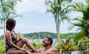 Um casal relaxando à beira de uma piscina, cercado por vegetação exuberante e palmeiras.