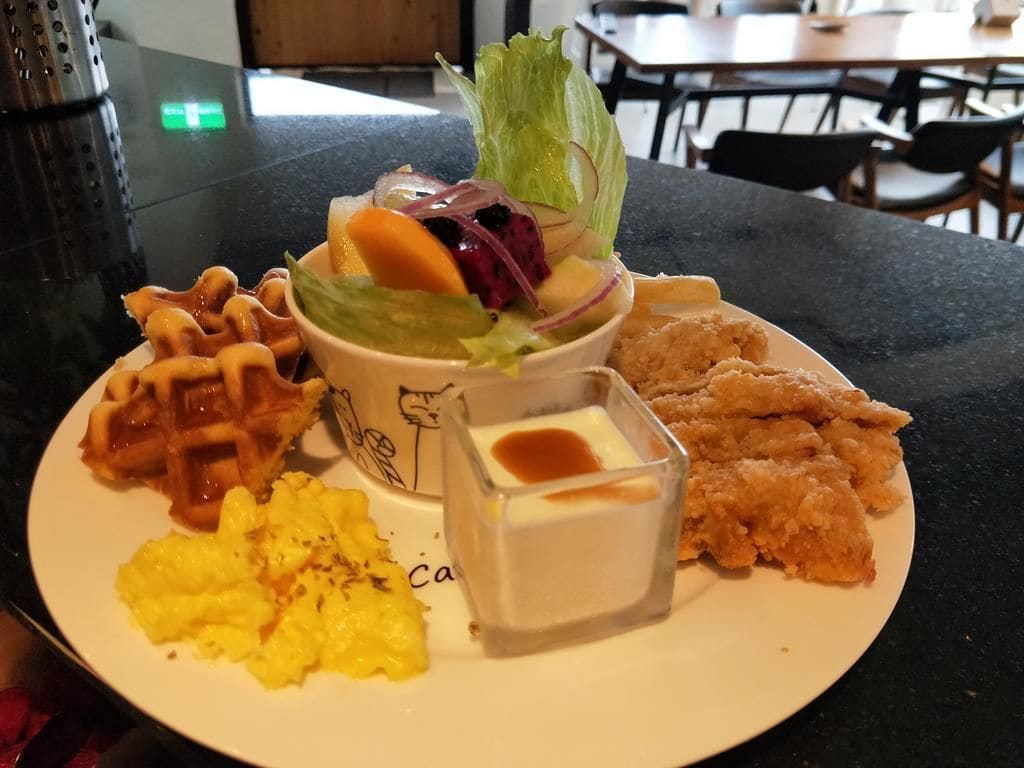 Prato de comida com waffles, ovos mexidos, frango frito e uma salada, acompanhado de um pequeno copo de leite.