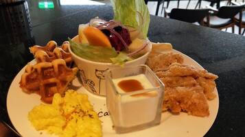 Prato de comida com waffles, ovos mexidos, frango frito e uma salada, acompanhado de um pequeno copo de leite.
