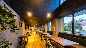 Um restaurante comprido e estreito com mesas de madeira e cabines verdes, janelas amplas e plantas em vasos ao longo das paredes.