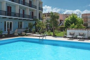 Uma piscina com escada, espreguiçadeiras e um edifício com varandas e janelas.